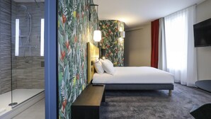 Quarto de hotel com chuveiro, cama, um banquinho, televisão e parede com papel de parede tropical.