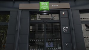 Entrada de hotel com toldo de vidro, placa indicando "ibis Styles Hotels" e o número 97 no edifício.