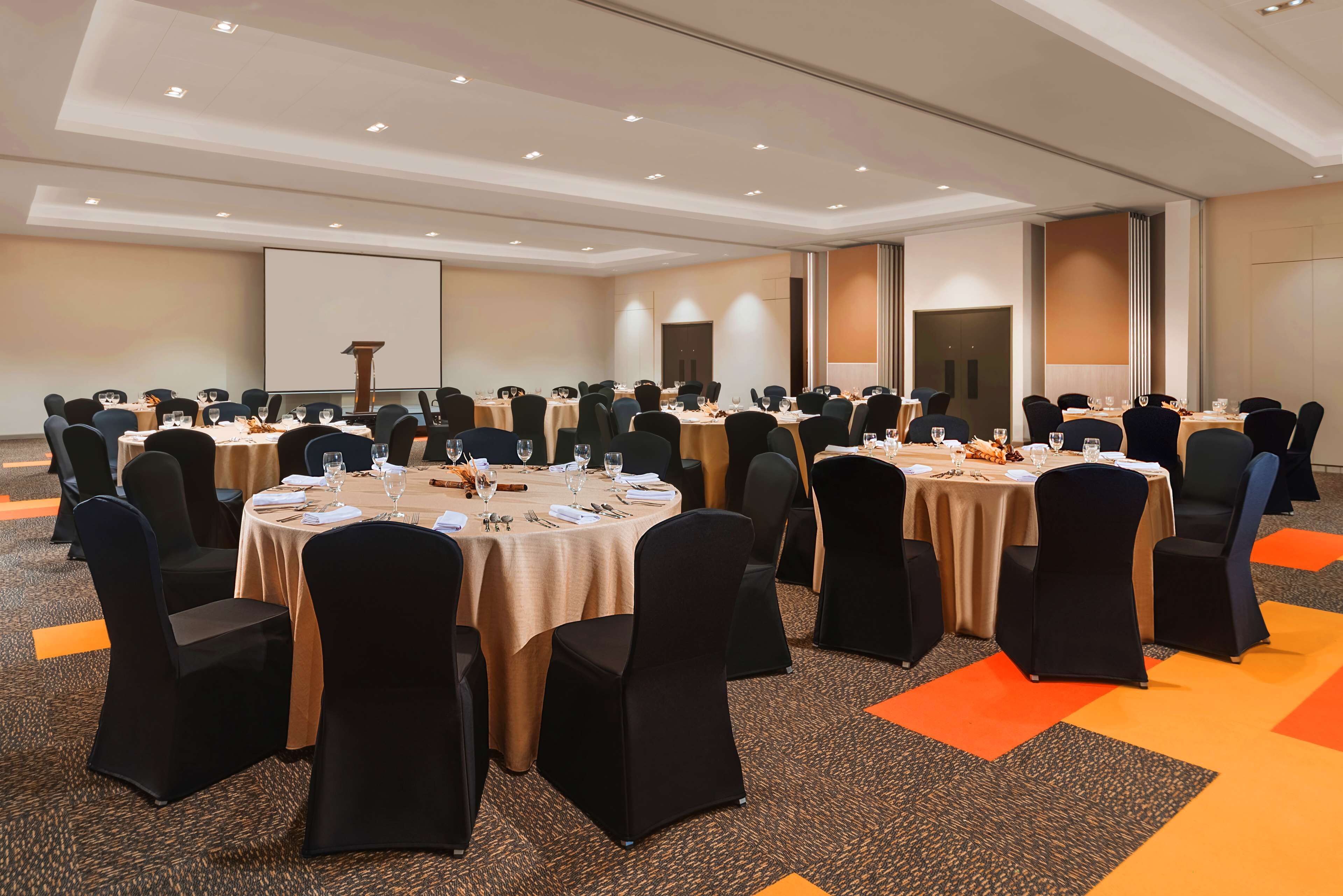 Uma sala de conferências com várias mesas redondas, cadeiras pretas e toalhas de mesa bege.