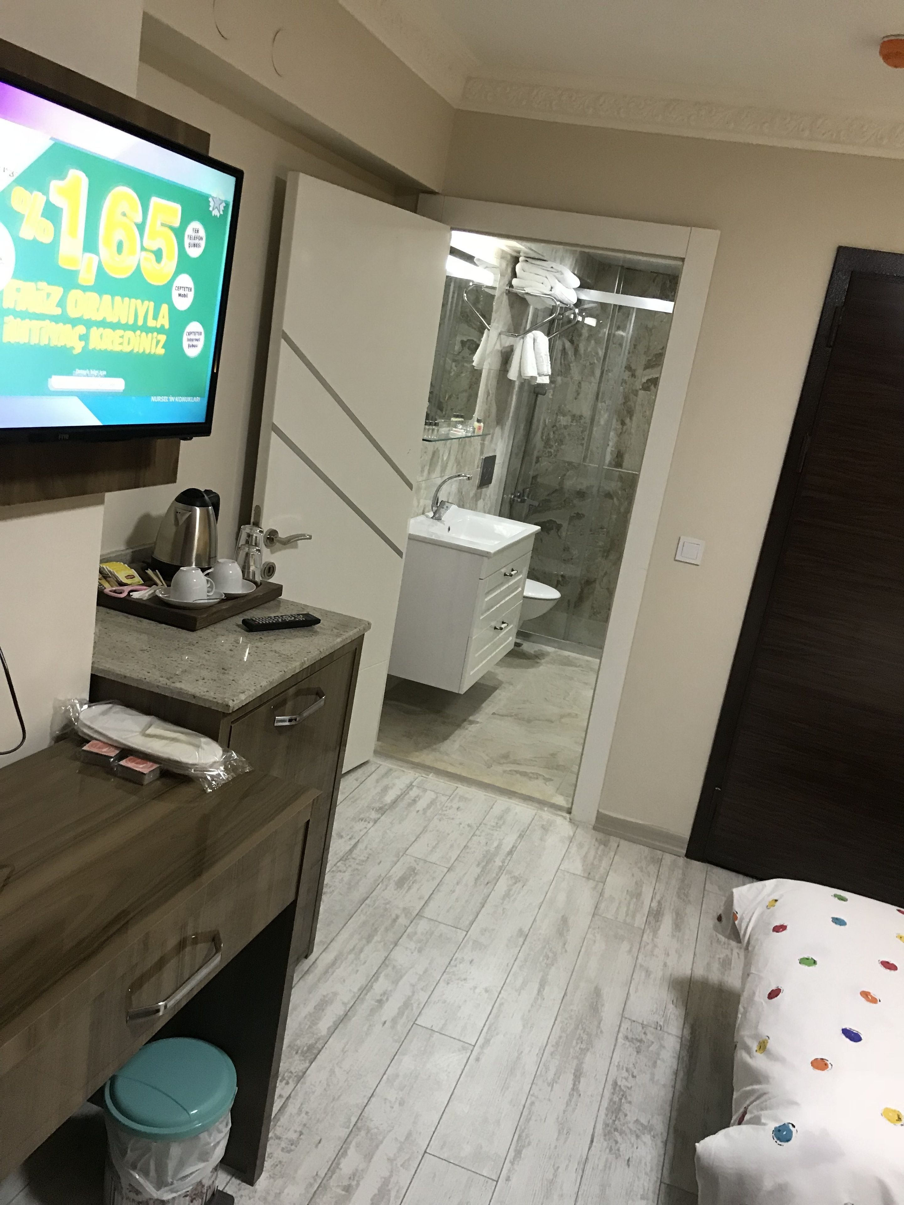 Quarto de hotel com televisão exibindo uma promoção, uma escrivaninha de madeira com chaleira, um cesto de lixo e uma cama com travesseiro estampado em poá.