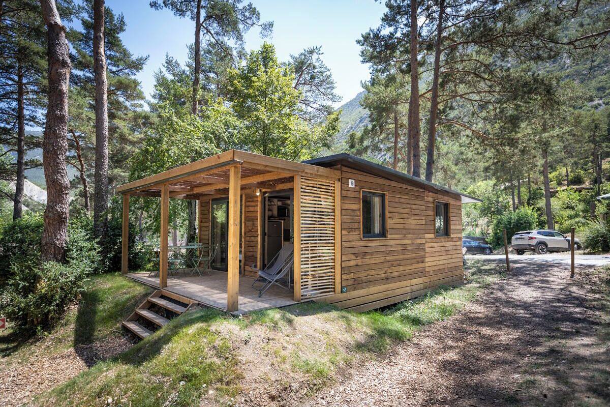 Uma cabana de madeira com varanda coberta, um pequeno alpendre e uma construção térrea rodeada por árvores.