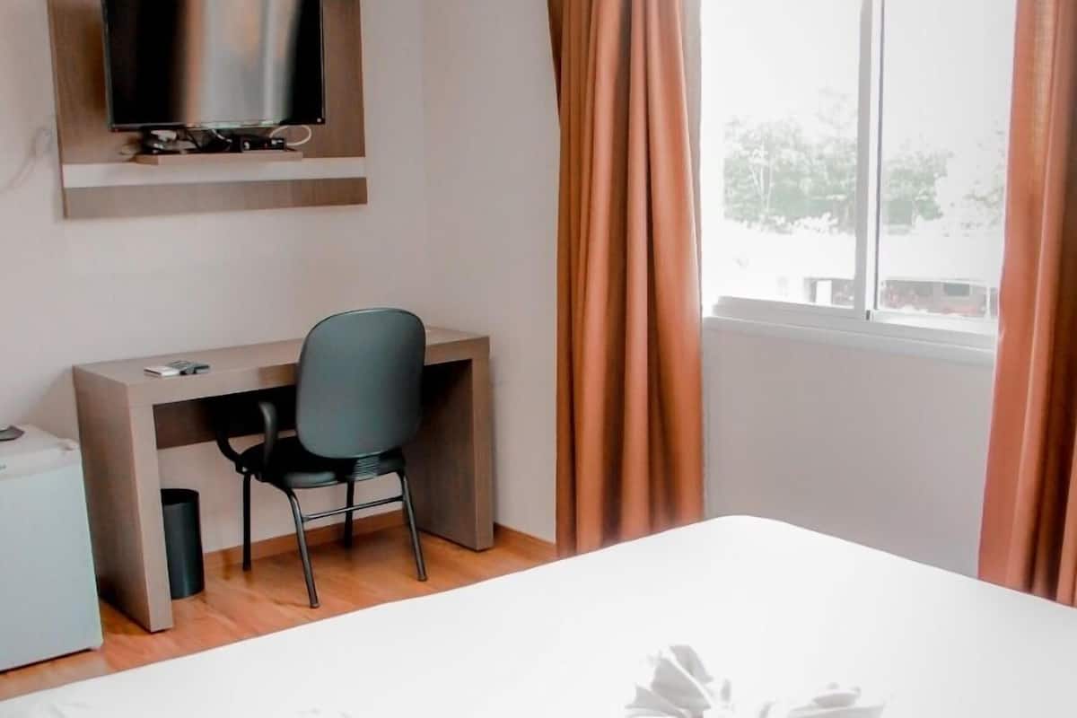 Quarto de hotel com cama, escrivaninha, cadeira, televisão e janela com cortinas.