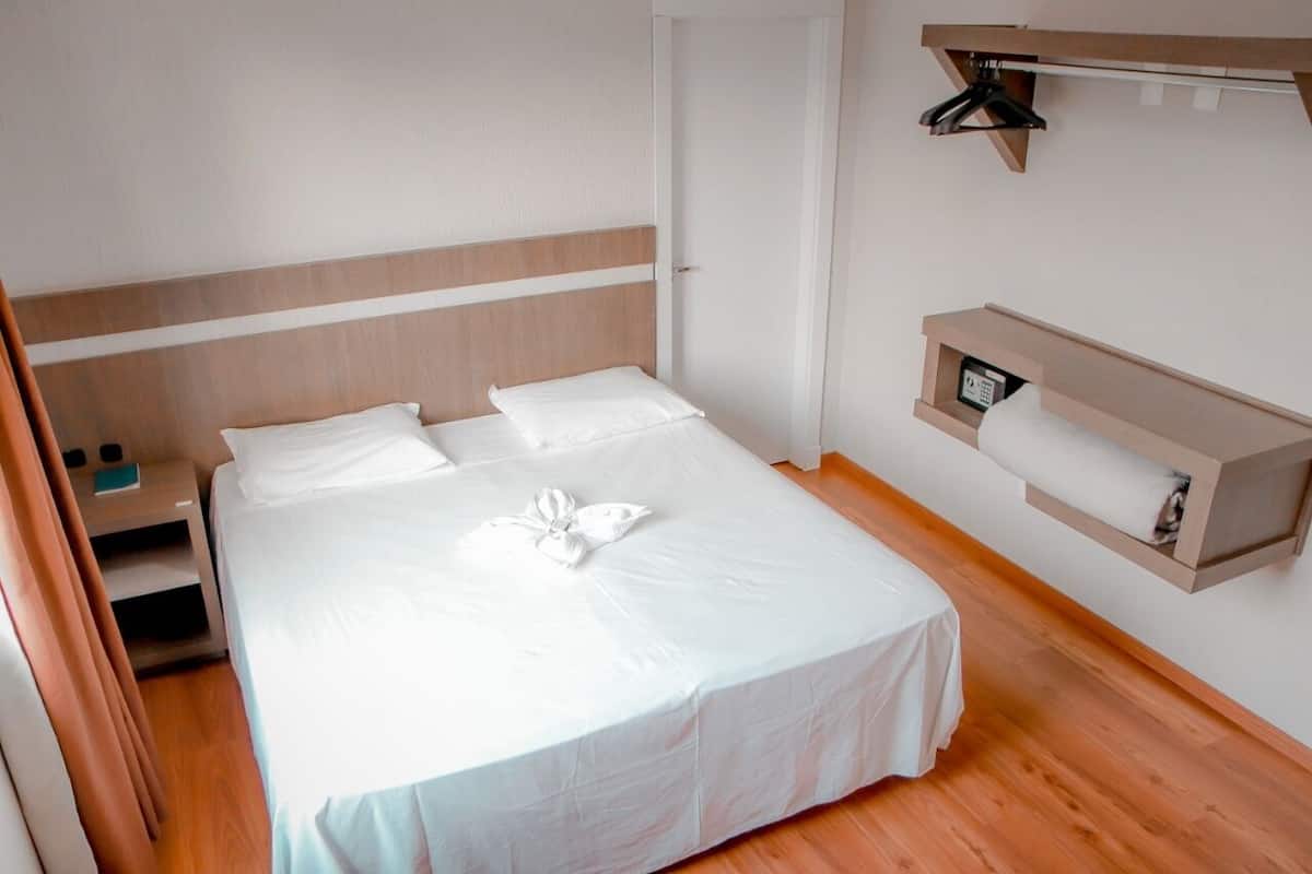 Cama bem arrumada com colcha e travesseiros brancos, cabeceira de madeira, mesa de cabeceira com caixa de lenços e prateleira na parede com toalha e uma caixa pequena.