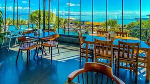 Restaurante com amplas janelas que oferecem vista para o mar e uma ponte. Mesas e cadeiras dispostas para refeições.