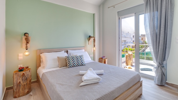 Quarto moderno com uma cama grande, uma mesinha de cabeceira de madeira, uma pequena mesa com abajur e vista para a cidade através de uma porta deslizante.