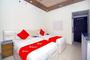 Quarto de hotel com duas camas, teto de madeira, parede branca, uma porta e uma pequena escrivaninha.