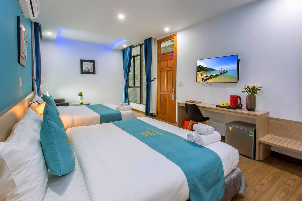 Quarto de hotel com duas camas, uma escrivaninha, uma televisão e um frigorífico pequeno.