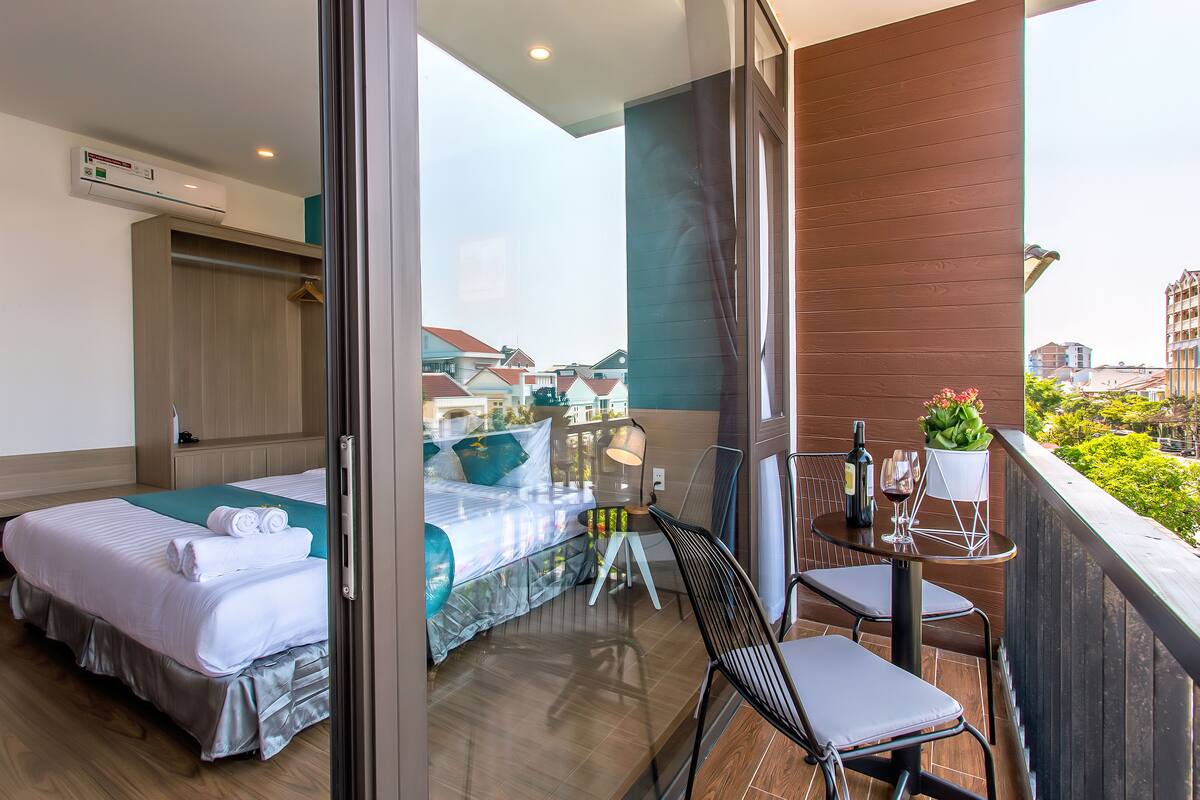 Quarto de hotel com duas camas, varanda com mesa e cadeiras, e vista da cidade.