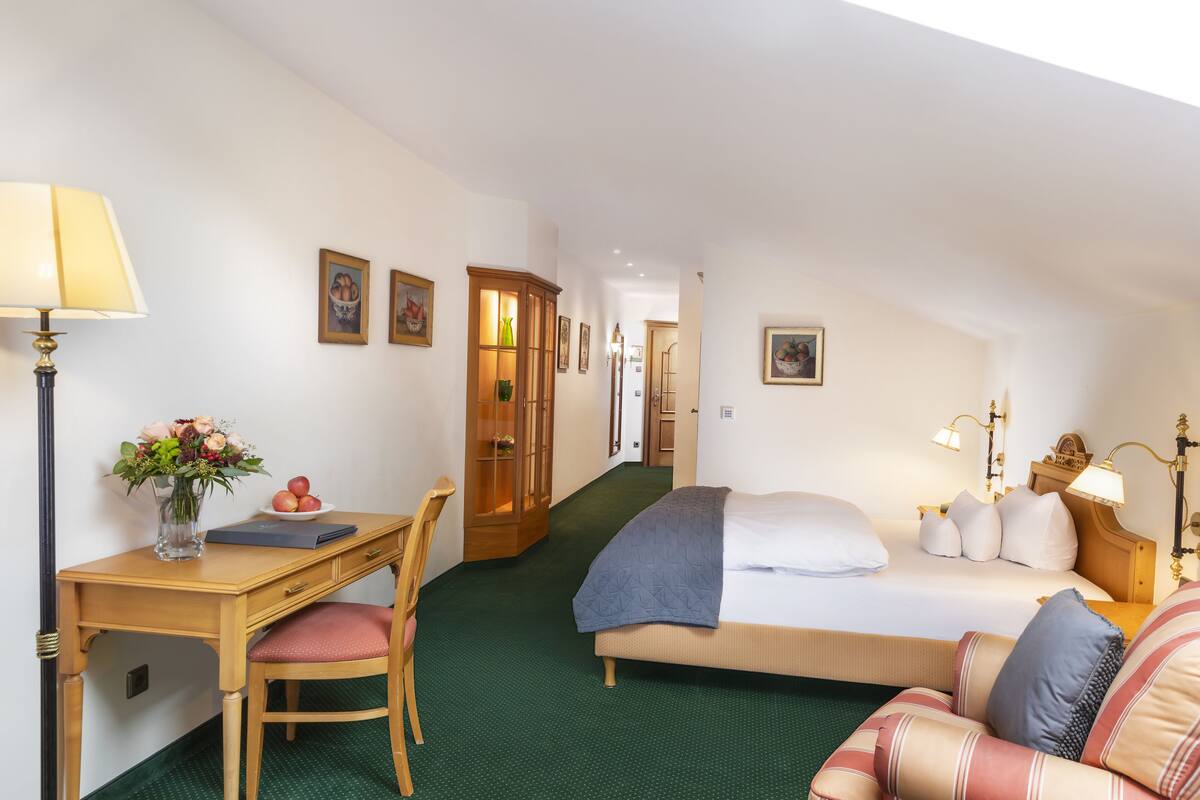 Quarto de hotel com cama, escrivaninha, cadeira, luminária e um vaso com flores.