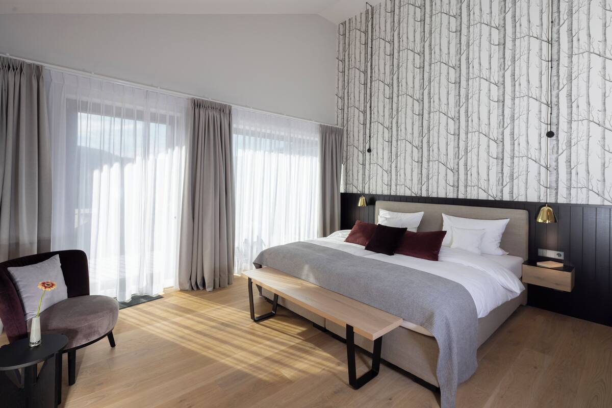 Quarto de hotel moderno com uma cama grande, um banco, uma cadeira e uma mesa de cabeceira.