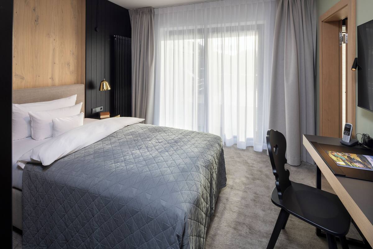 Quarto de hotel com cama grande, escrivaninha com cadeira, televisão e janela com cortinas.