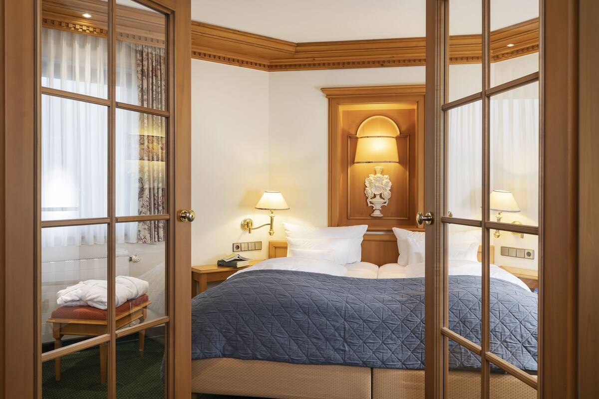 Quarto de hotel com cama grande, duas mesas de cabeceira, um criado-mudo, um espelho e uma janela com cortinas.