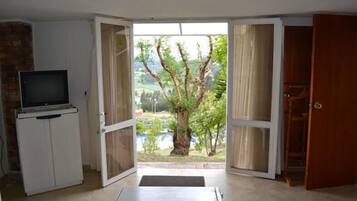 Quarto com televisão sobre um armário, porta de vidro deslizante para um jardim com árvore e piscina.