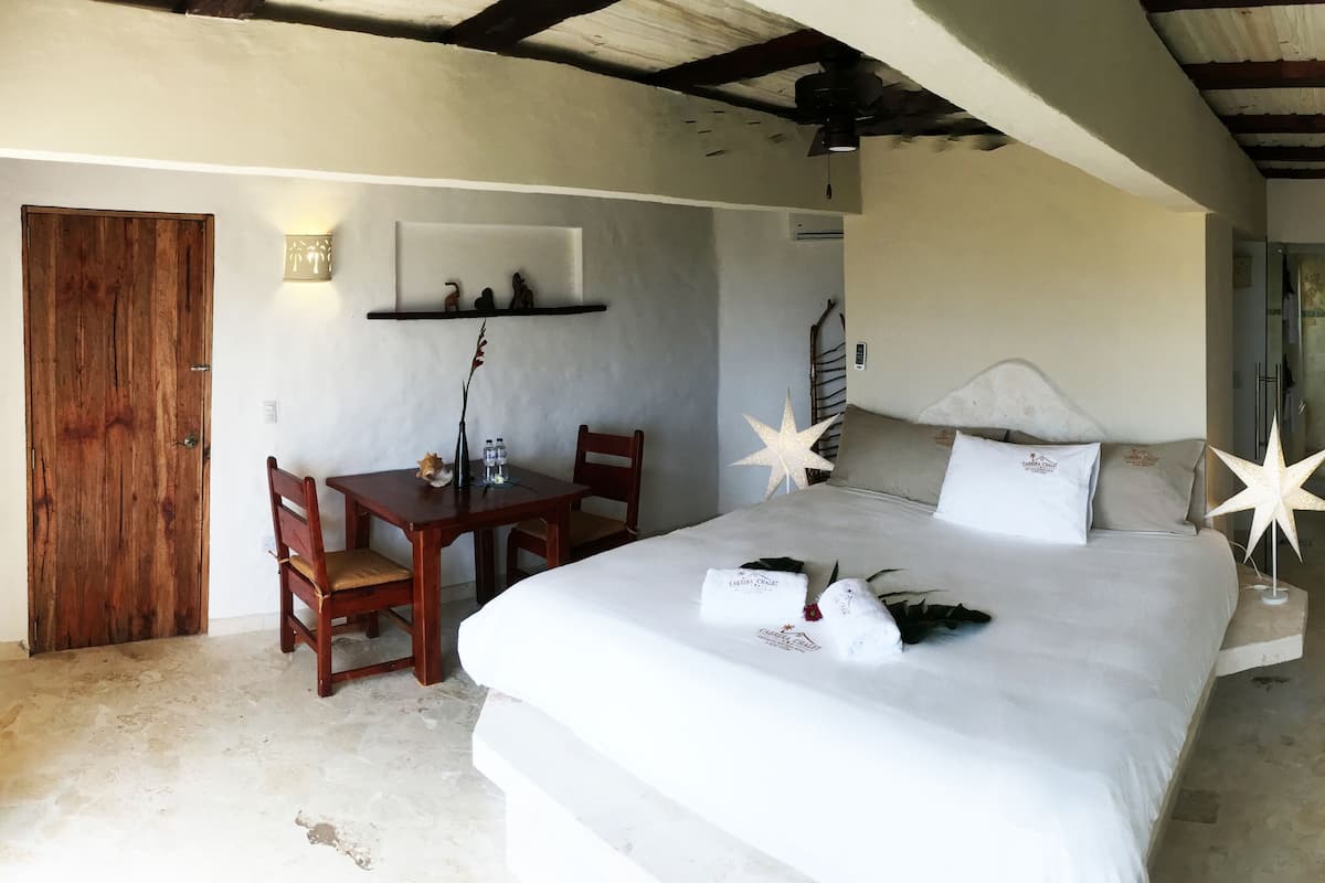 Quarto com cama, mesas de cabeceira, escrivaninha e uma cadeira. Há uma porta, uma estante com objetos decorativos e um vaso com planta.