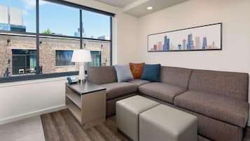 Sala de estar moderna com sofá seccional, uma mesinha com abajur e vista para a cidade através de amplas janelas.