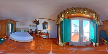 Quarto de hotel com uma cama grande, uma escrivaninha com cadeira, uma mesinha com um vaso de flores, uma luminária e uma janela ampla com cortinas.