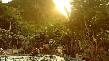 Piscina com uma pessoa na água, cercada por vegetação exuberante e uma colina ao fundo.