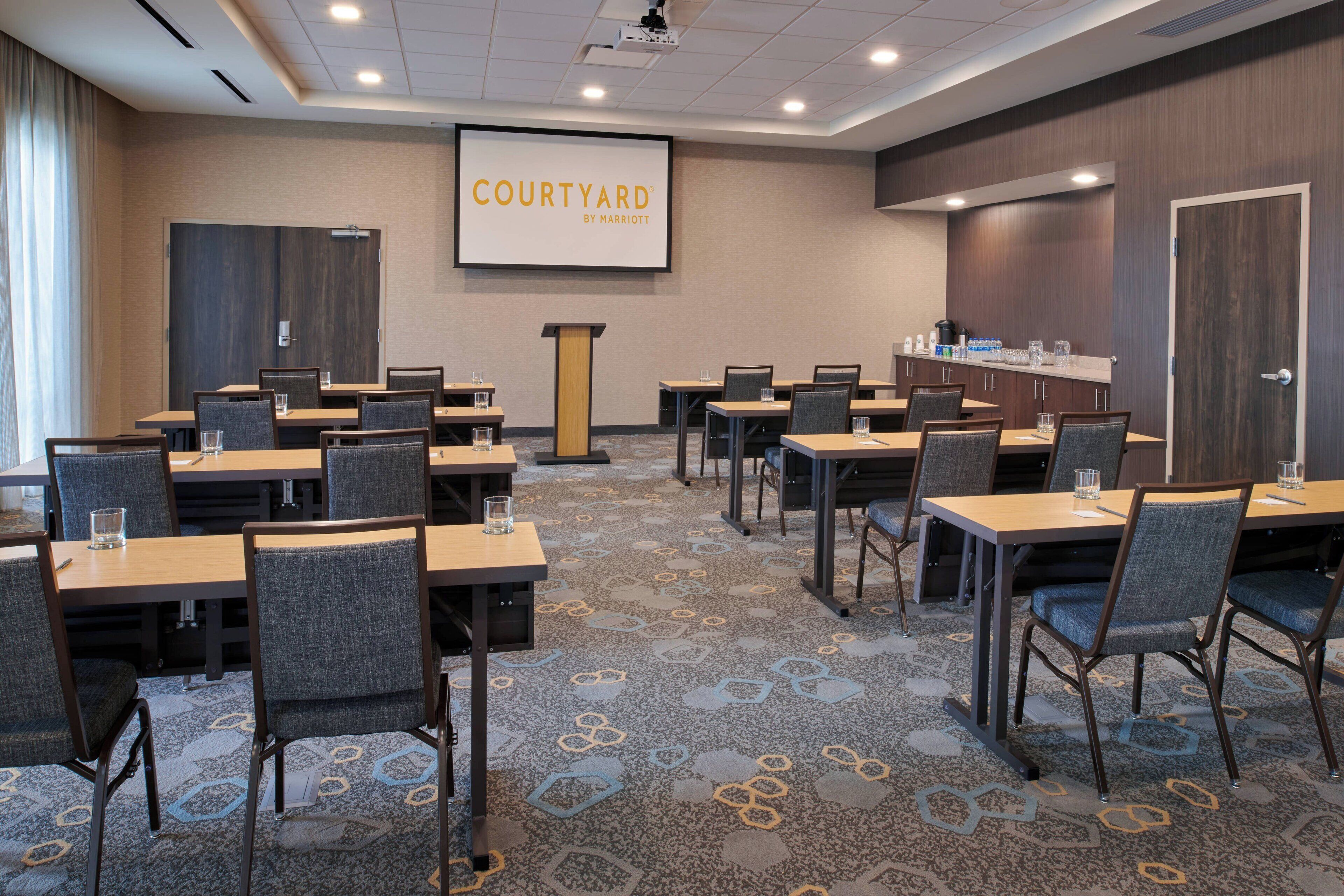 Uma sala de conferência com várias mesas e cadeiras, um púlpito e uma tela exibindo "COURTYARD BY MARRIOTT".