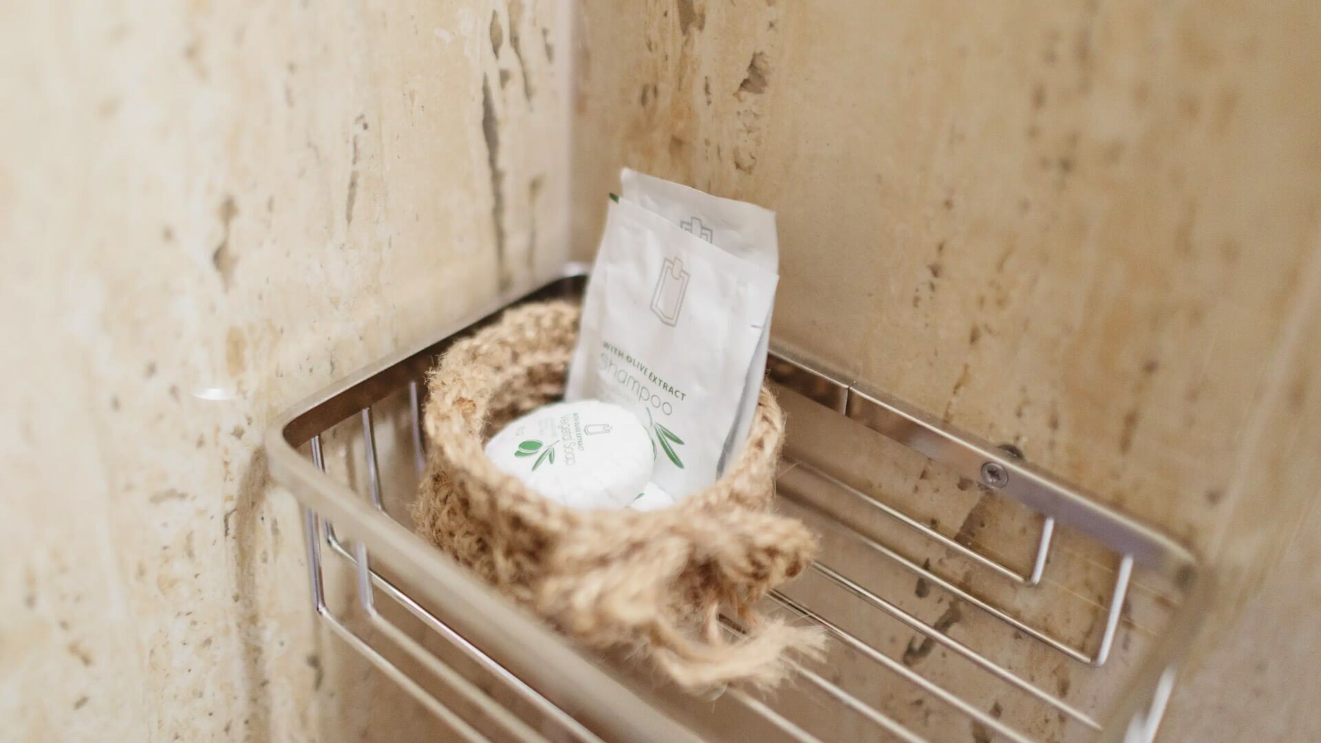 Uma prateleira de metal com uma cesta trançada contendo uma garrafa branca e uma embalagem branca com a inscrição 'EXTRATO DE OLIVA SHAMPOO'.