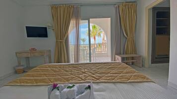 Quarto de hotel com uma cama grande, televisão na parede, escrivaninha e varanda com vista para palmeiras.