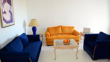 Sala de estar com dois sofás, um azul e um laranja, uma mesa de centro de vidro com frutas, um abajur e um laptop sobre uma mesinha lateral.