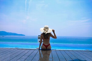 Uma pessoa de biquíni vermelho e chapéu branco sentada em um deck de madeira à beira de uma piscina, com vista para o oceano.