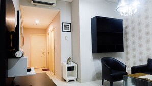 Uma sala de estar moderna com sofá de couro preto, mesa de centro de vidro e TV fixada na parede. Há uma mesinha de cabeceira branca com um objeto decorativo. O ambiente possui um lustre e papel de parede estampado.