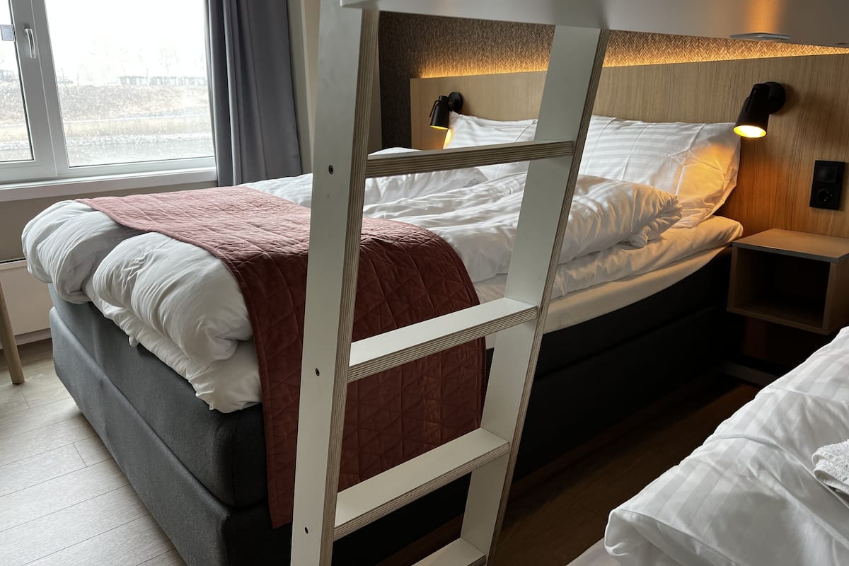 Quarto com beliche, cabeceira de madeira, mesas de cabeceira com luminárias e janela com cortinas.