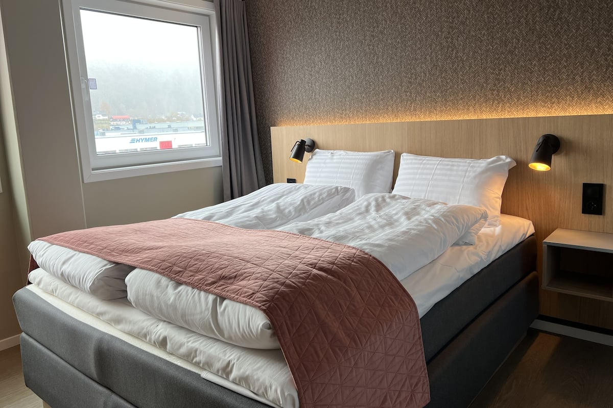 Cama bem arrumada com colcha rosa, travesseiros brancos e cabeceira cinza. Há duas luminárias de leitura fixadas na parede, de cada lado da cama.
