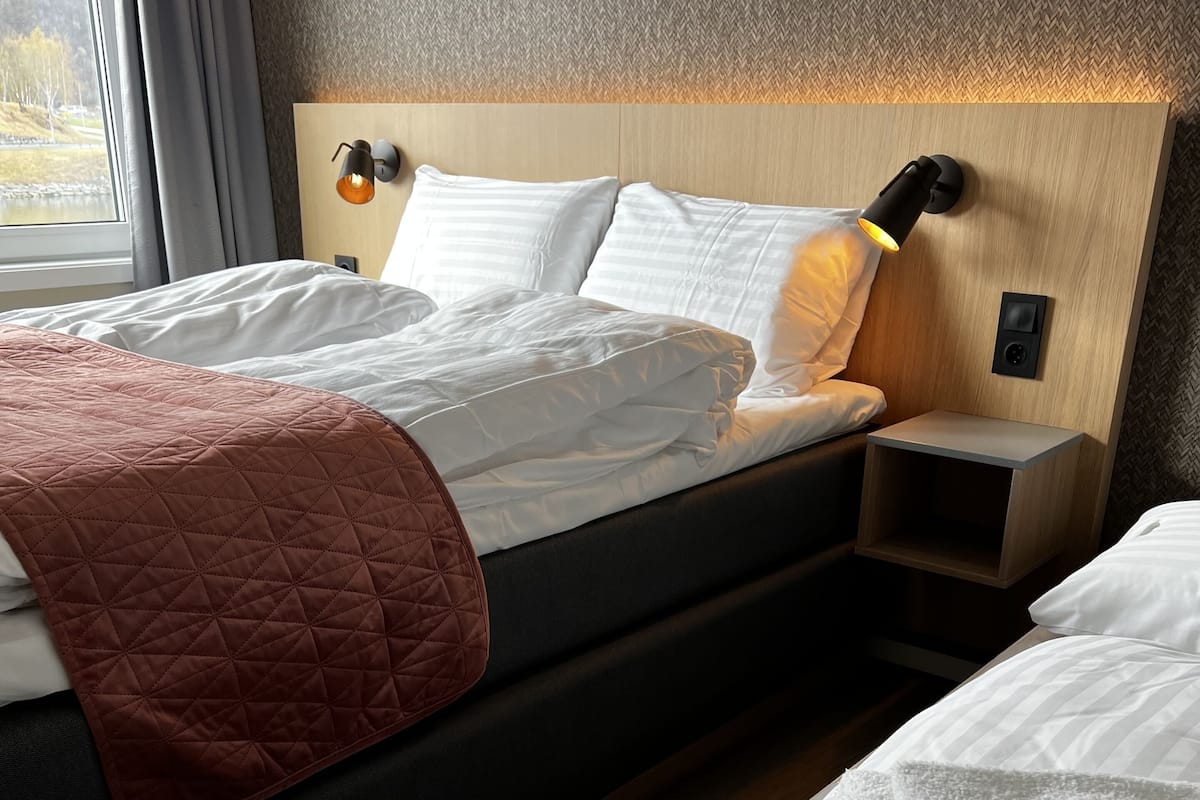 Uma cama de casal com colcha marrom, uma mesa de cabeceira com abajur e vista para um lago pela janela.
