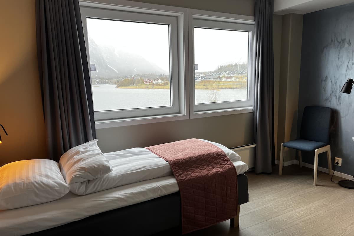 Quarto com cama, uma cadeira e vista para um lago e montanhas através de grandes janelas.