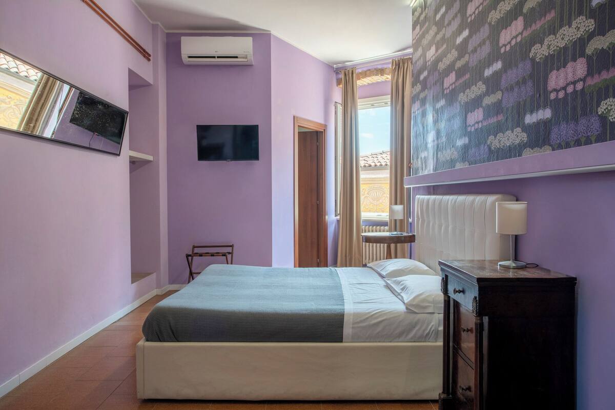 Quarto com cama, televisão, ar condicionado, janela com cortinas e parede com padrão floral.