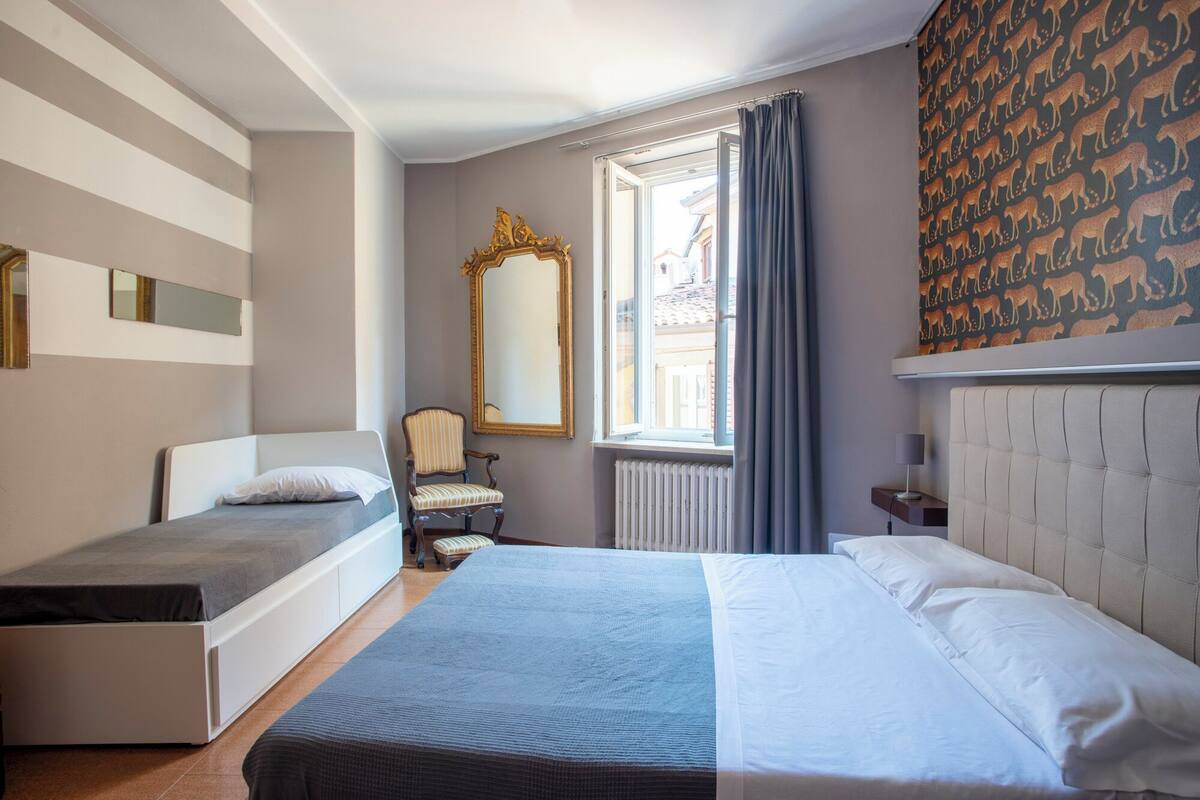 Quarto de hotel com duas camas, janela com cortinas, radiador, uma cadeira, um espelho e parede estampada.