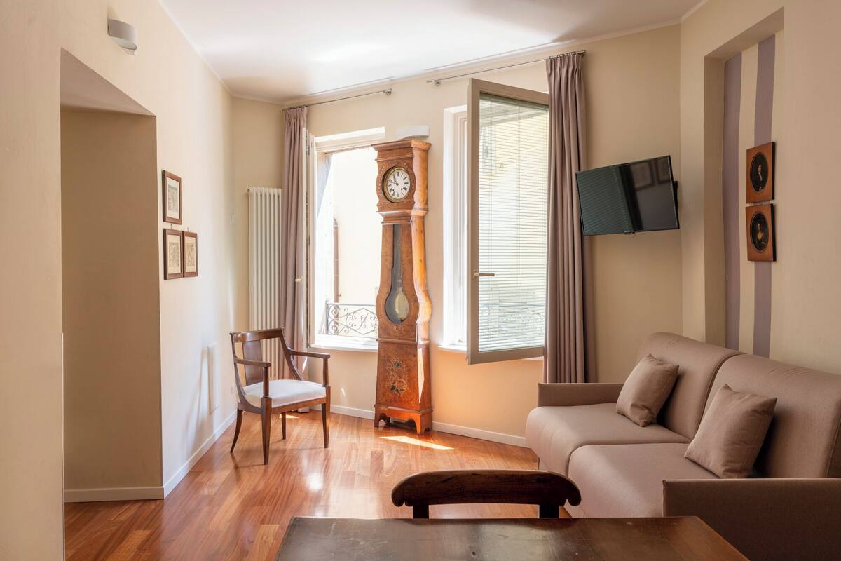 Uma sala de estar com piso de madeira, um sofá, uma poltrona, uma mesa, um relógio, uma TV e uma janela com cortinas.