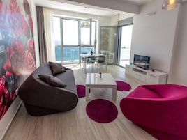 Sala moderna com sofá, mesa de centro e televisão. Varanda com vista para o mar.
