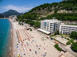 Uma praia com fileiras de guarda-sóis e espreguiçadeiras, um prédio grande com vários andares e montanhas ao fundo.