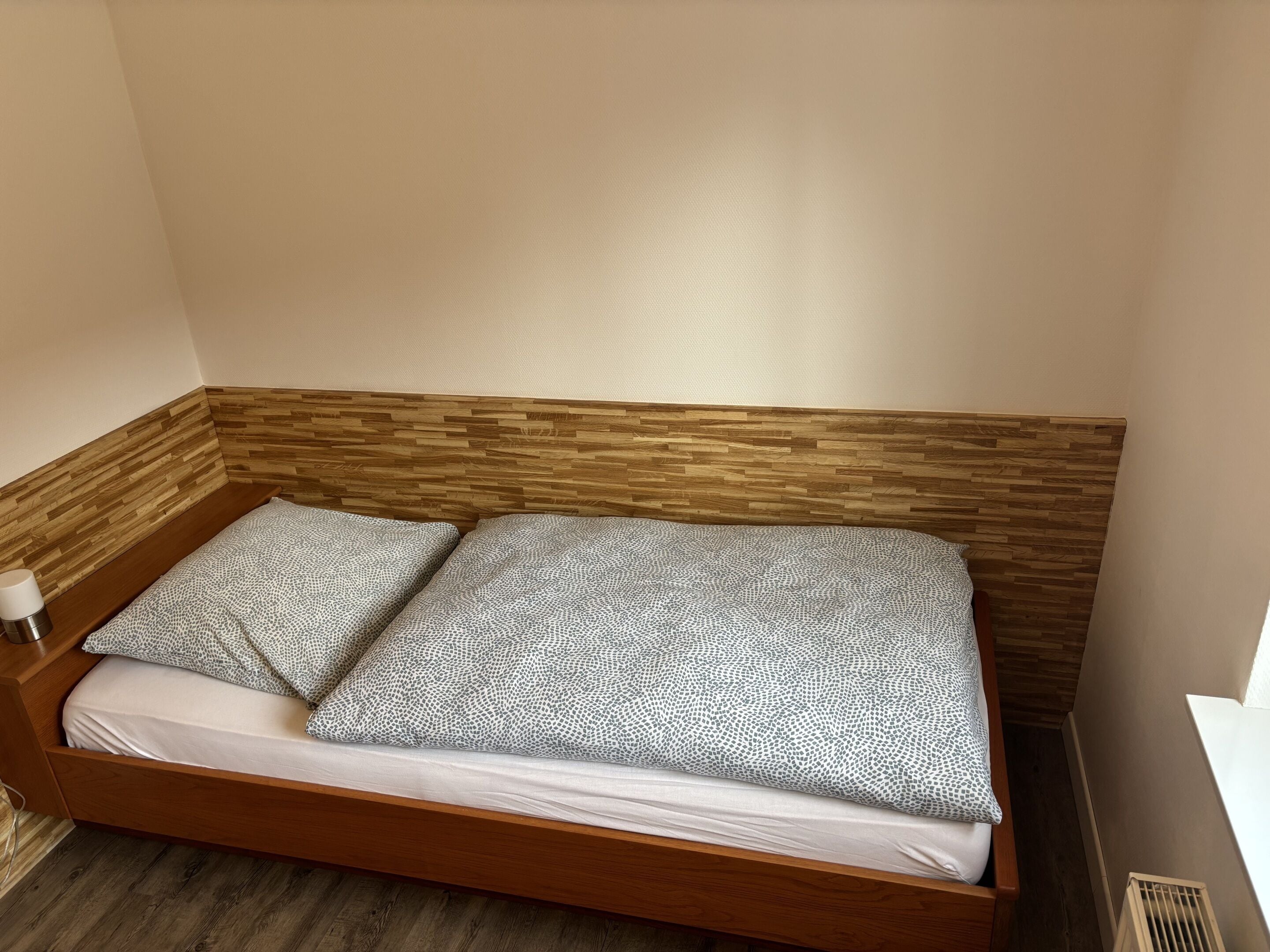 Quarto pequeno com uma cama de solteiro, cabeceira de madeira e mesa de cabeceira.