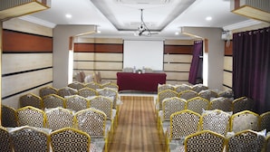 Uma sala de conferências com fileiras de cadeiras estampadas, uma mesa vermelha com tela de projetor e um ventilador de teto.