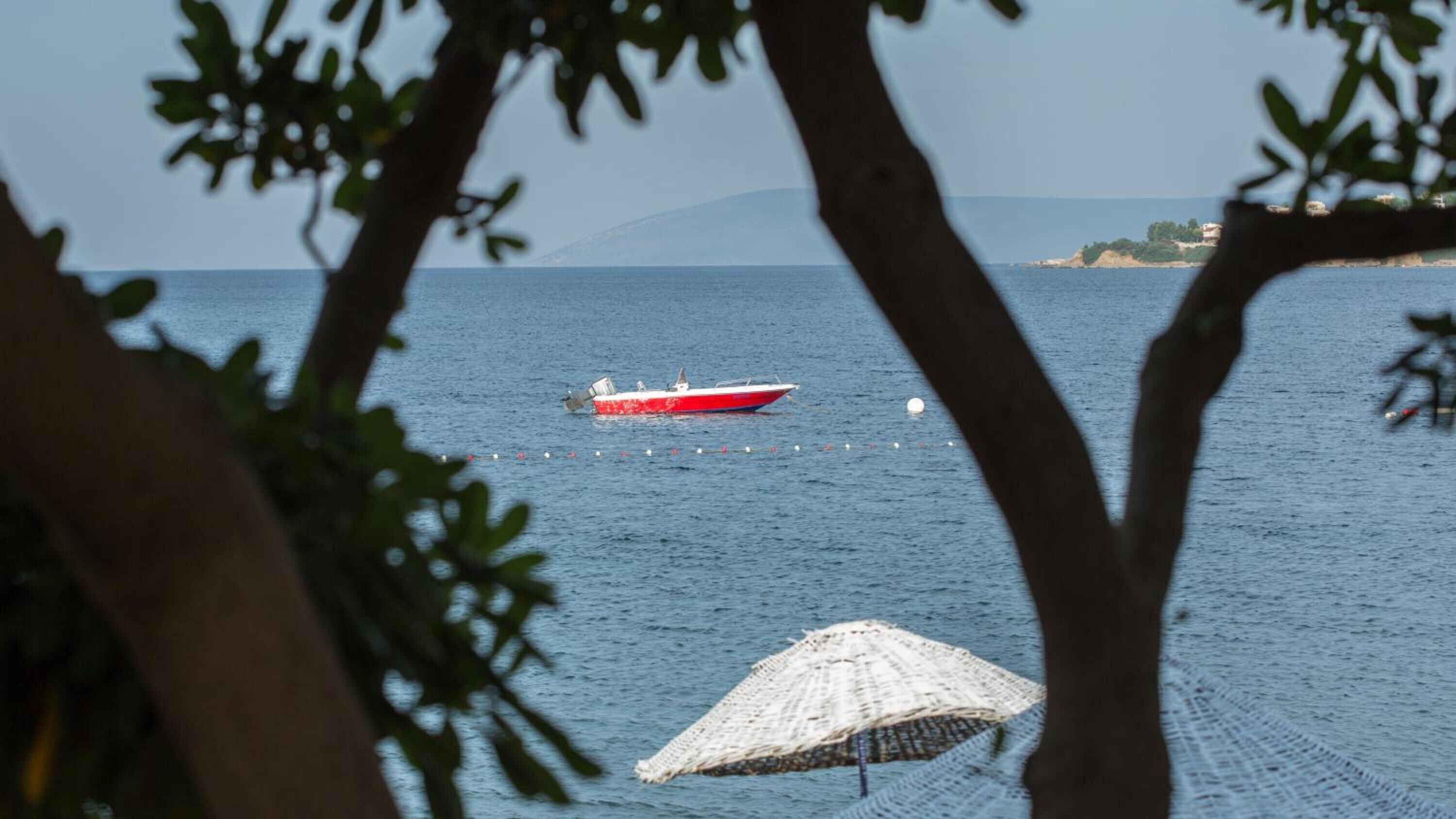 Um barco em águas calmas, um guarda-sol de palha e uma linha costeira distante com edifícios.
