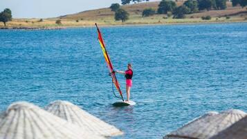 Uma pessoa praticando windsurf em um corpo de água, com céu claro e colinas ao longe.