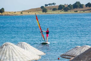 Uma pessoa praticando windsurf em um corpo de água, com céu claro e colinas ao longe.