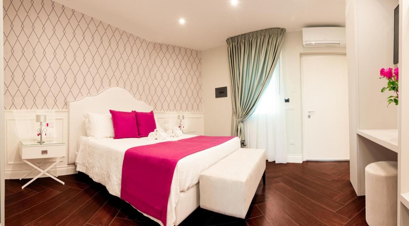 Quarto de hotel com cama branca, uma mesinha de cabeceira branca, mesa de cabeceira com abajur, janela com cortinas e ar-condicionado fixado na parede.