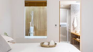 Um quarto moderno com uma cama grande, uma janela com persianas, uma mesa de cabeceira com um porta-cosméticos e um banheiro visível por uma porta aberta.