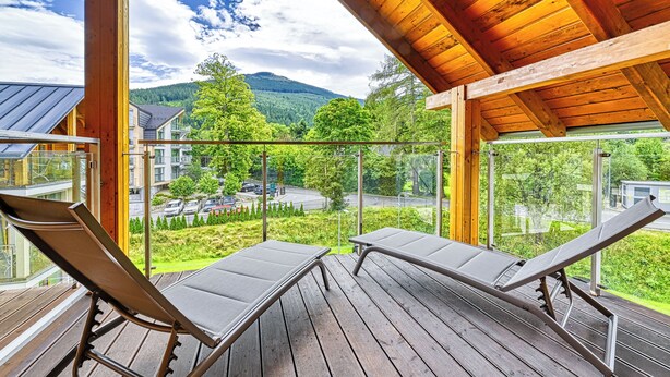 Um deck de madeira com duas espreguiçadeiras, com vista para uma área residencial e uma montanha.