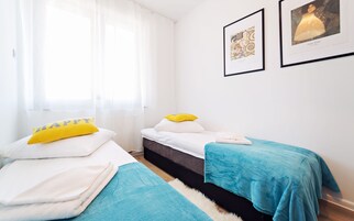 Quarto com duas camas, cada uma com um cobertor azul e um travesseiro amarelo. Há duas imagens emolduradas na parede.