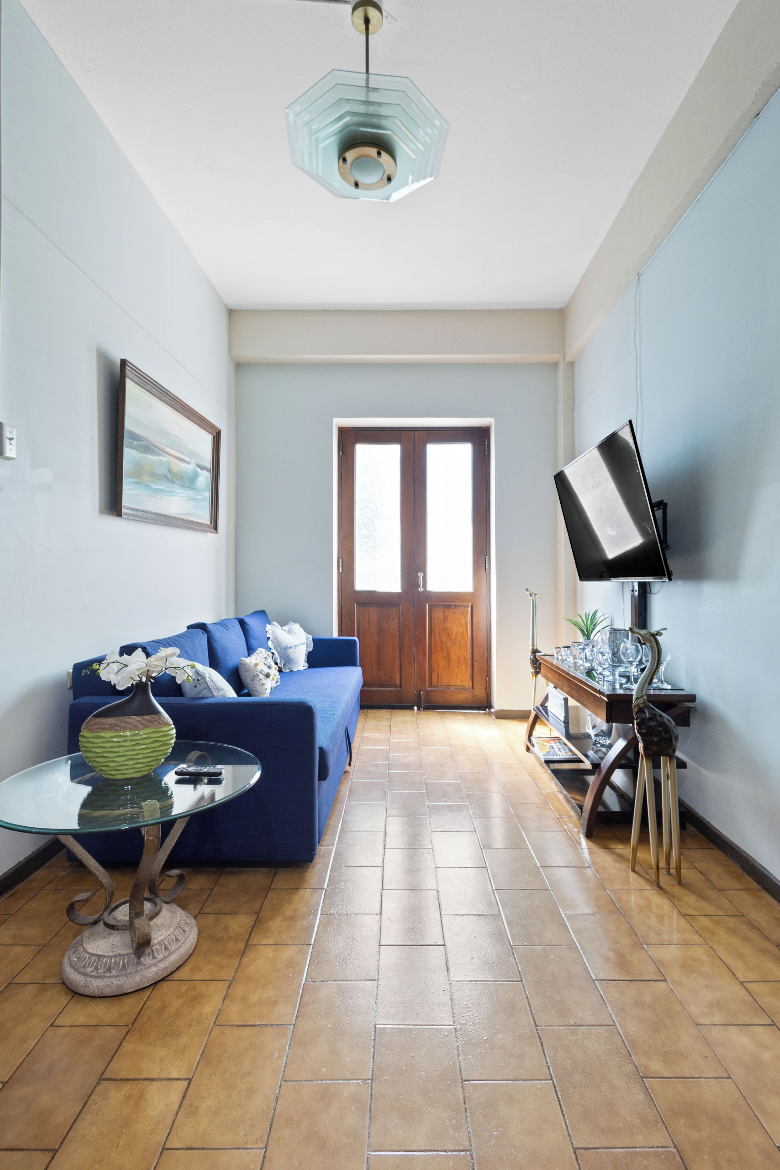 Uma sala de estar com um sofá azul, uma mesa de centro de vidro, um quadro na parede e uma televisão sobre um suporte.