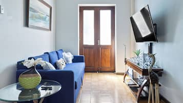 Uma sala de estar com um sofá azul, uma mesa de centro de vidro, um quadro na parede e uma televisão sobre um suporte.