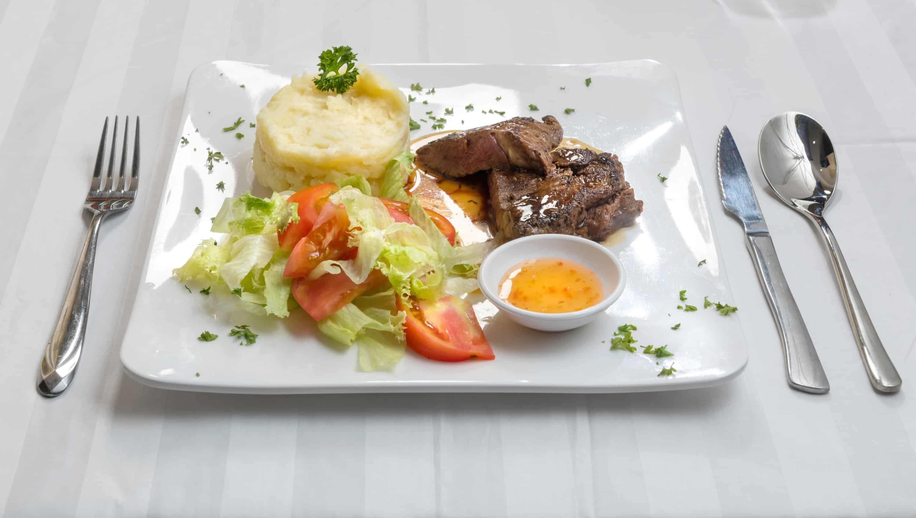 Um prato com purê de batatas, bife, salada e uma pequena tigela de molho.