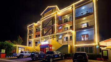 Um hotel com vários andares, varandas, uma escadaria central e um estacionamento com diversos veículos.