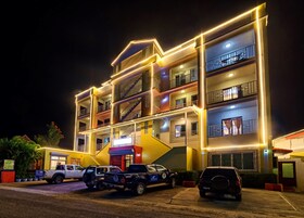 Um hotel com vários andares, varandas, uma escadaria central e um estacionamento com diversos veículos.